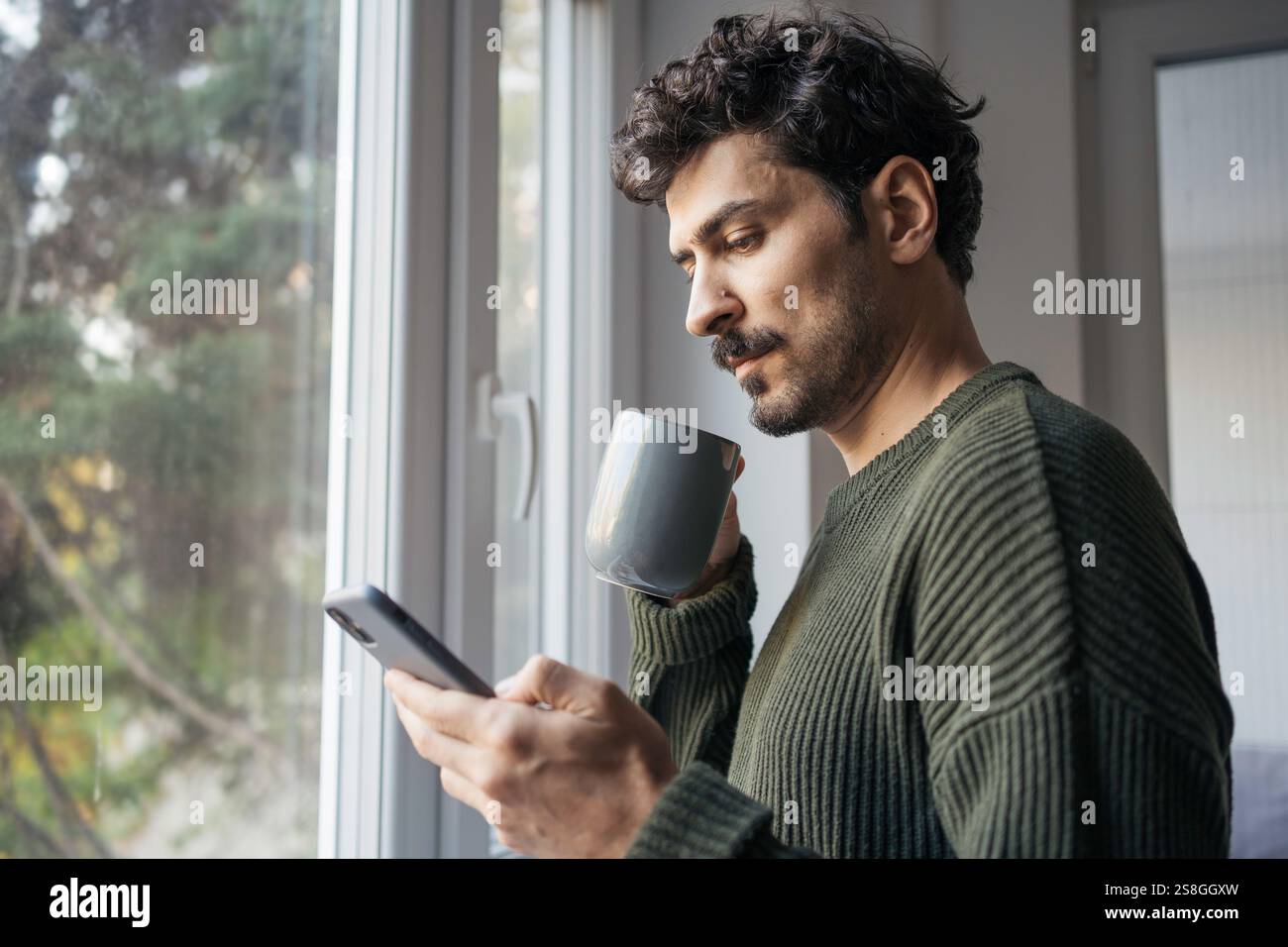 Portrait of young handsome middle eastern man scrolling on mobile phone in the morning standing ...