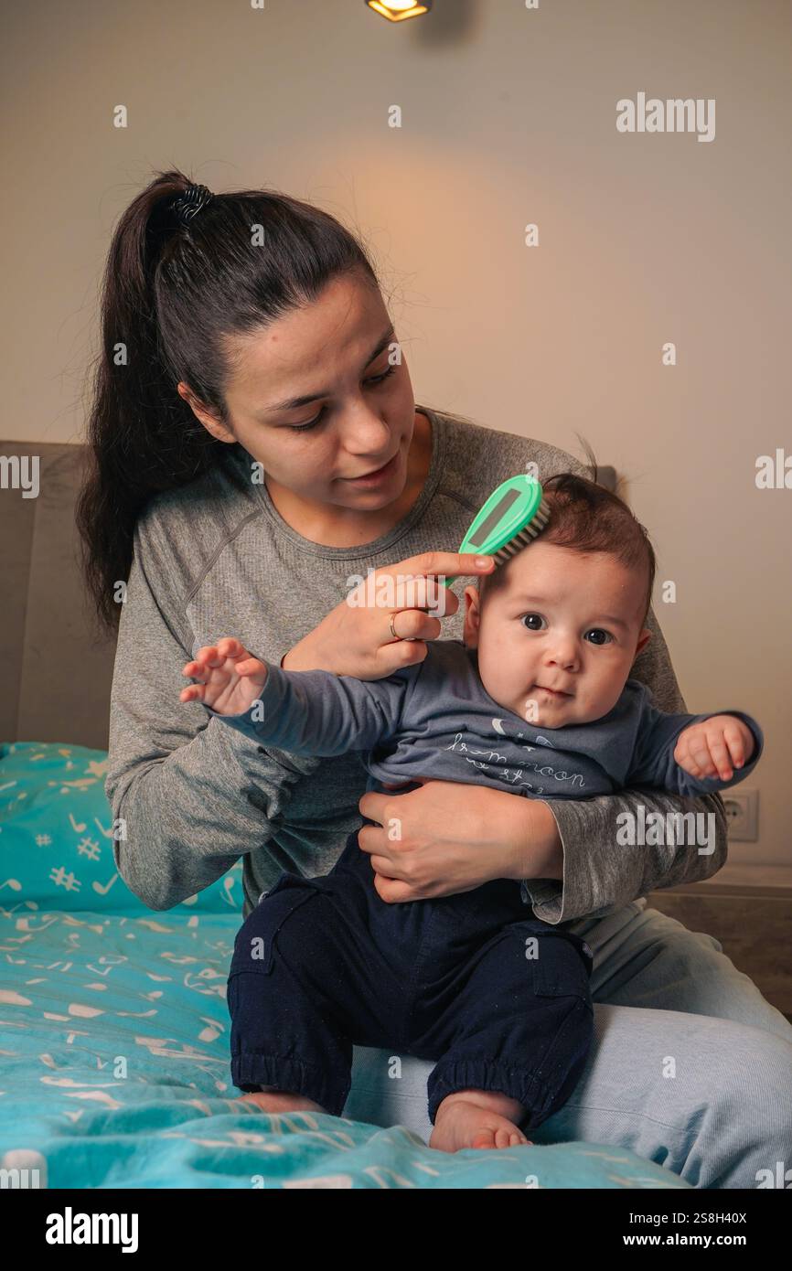 Young smiling mother combing the baby's hair with bristle brush. Taking ...