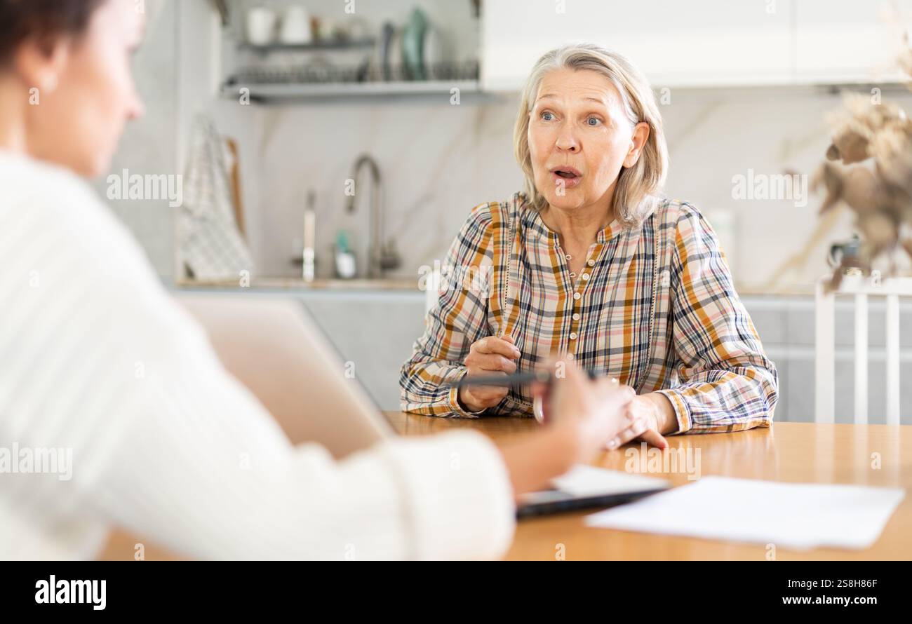Positive old woman sitting at the kitchen-table face to face with ...