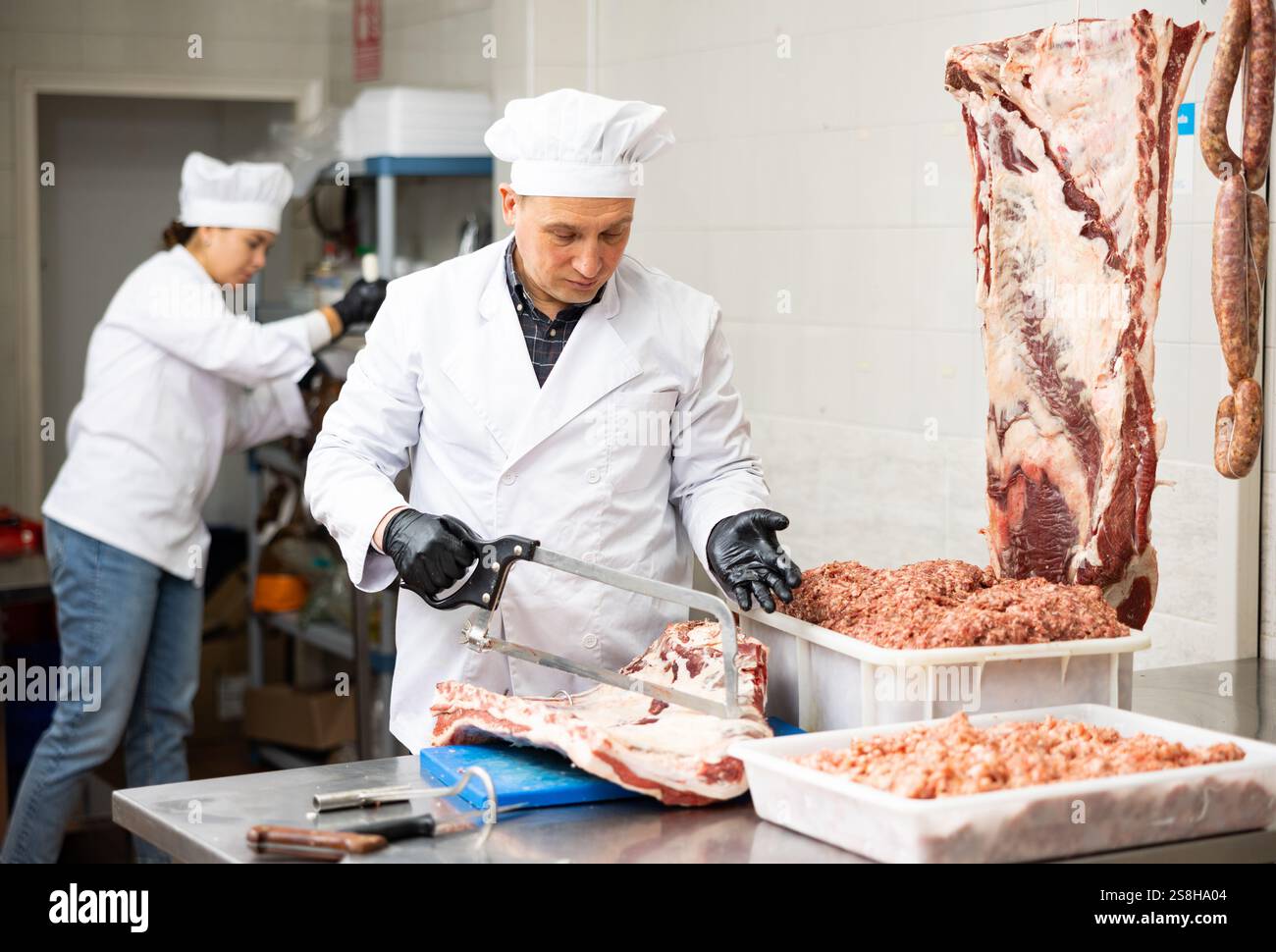 Smiling male butcher sawing large chunk of beef meat in butcher shop ...