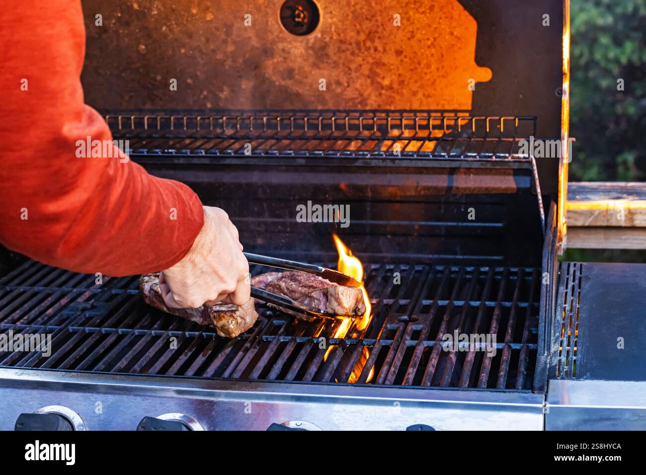 A person wearing a red sweater uses tongs to turn over juicy steaks on ...