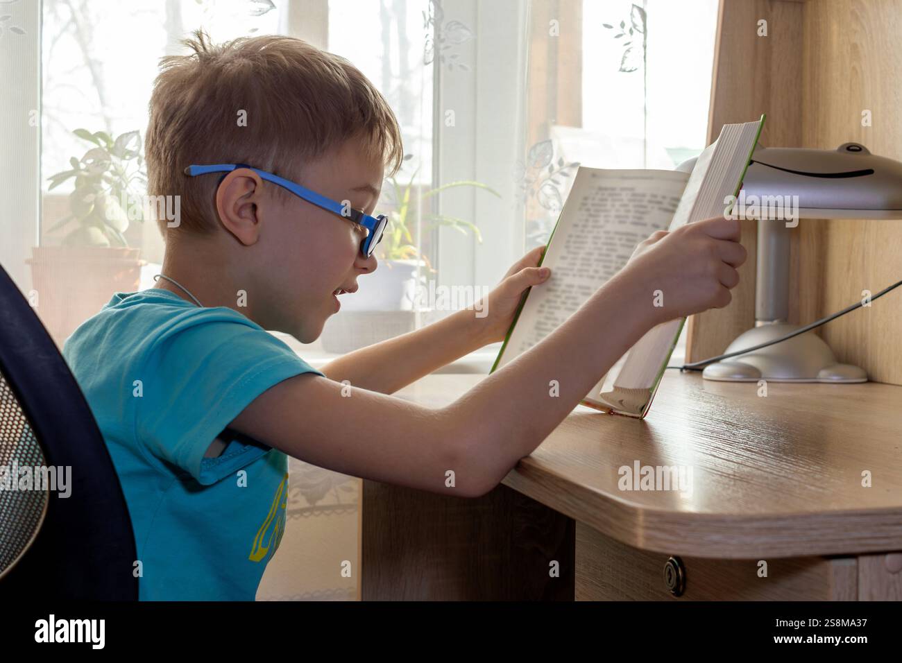 Boy with glasses enjoys reading book aloud at table by window with ...
