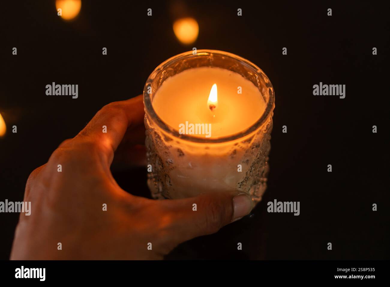 A hand holding a lit candle in a glass holder, with a dark reflective ...