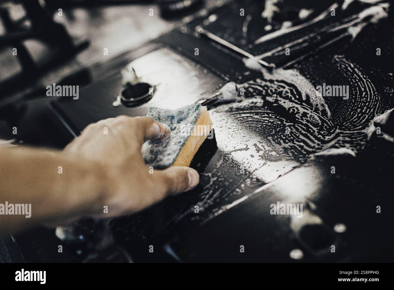 A hand cleaning a black stove with a soapy sponge in a domestic kitchen ...