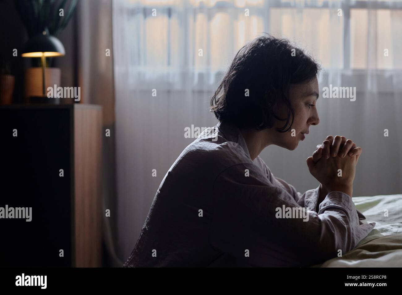 Side view shot of woman sitting still with clasped hand near made bed ...