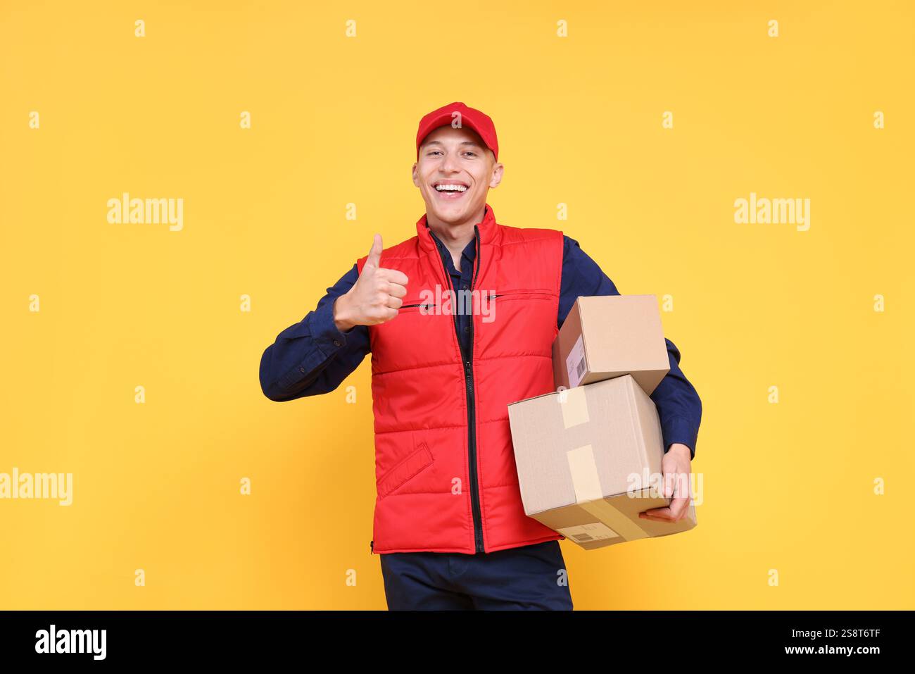 Happy postman with parcels showing thumbs up on yellow background Stock ...