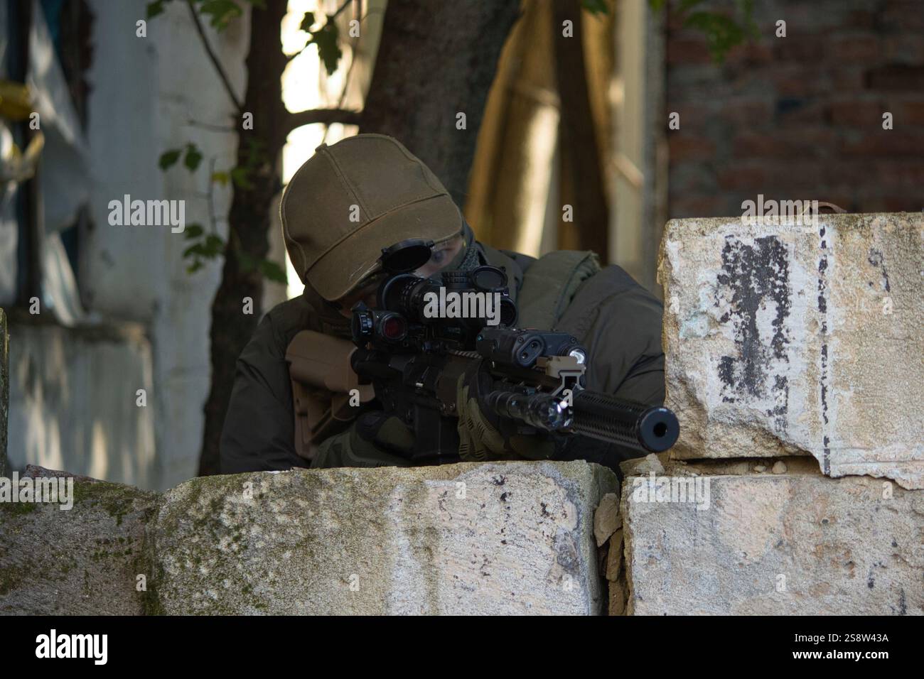 A tactical operator in military gear takes cover behind a moss-covered ...