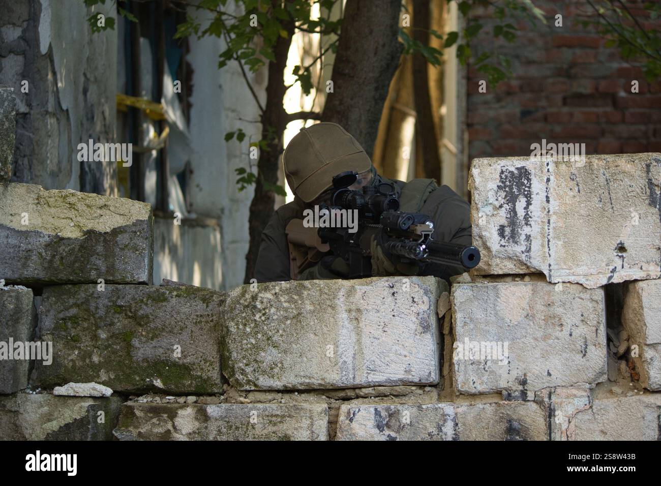 A tactical operator in military gear takes cover behind a moss-covered ...
