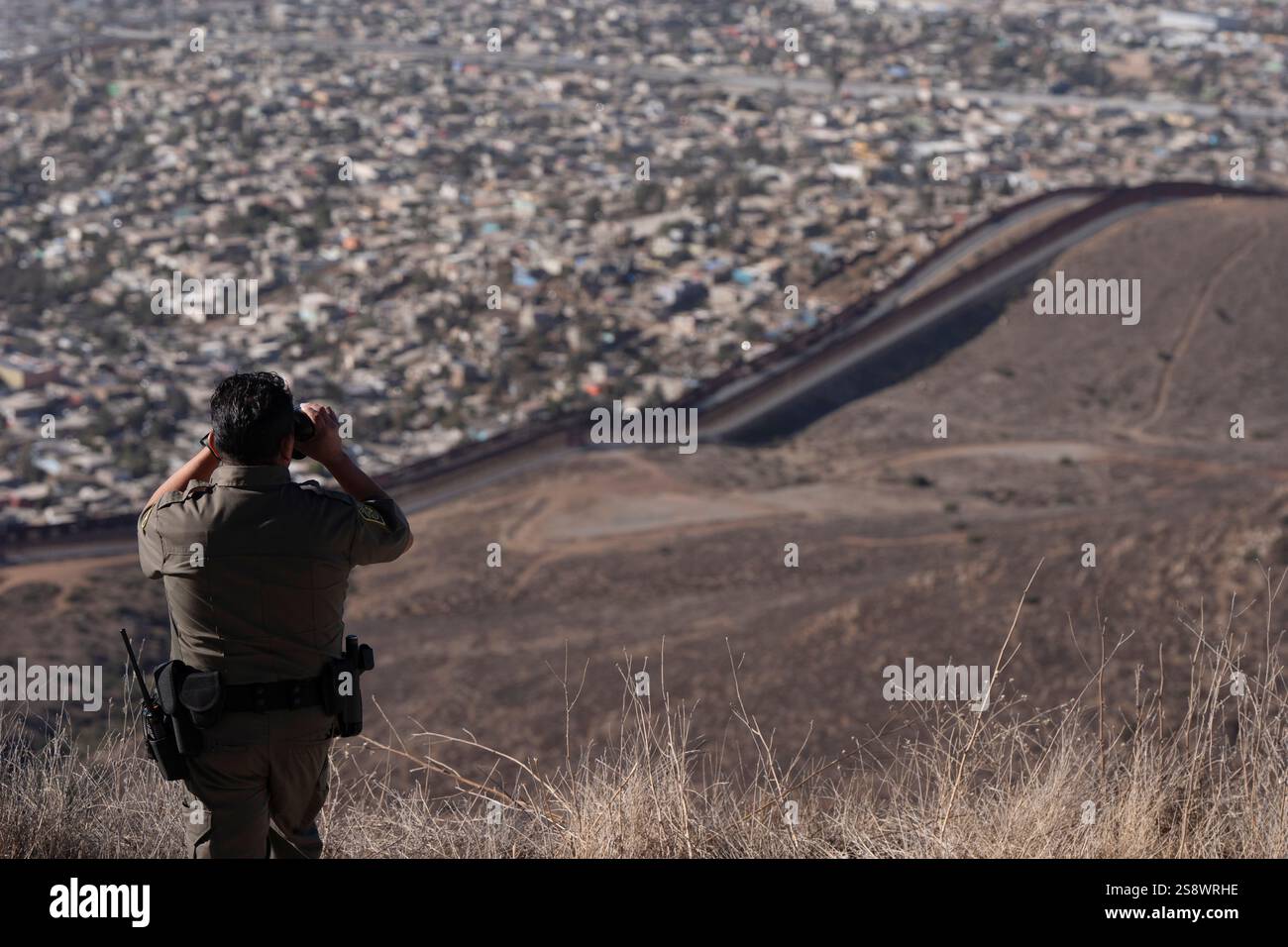 A Border Patrol agent looks through binoculars towards two border walls ...