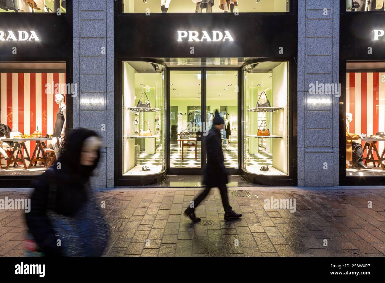Lugano, Switzerland - Dec 22, 2024: People walking past an illuminated Prada storefront in the ...