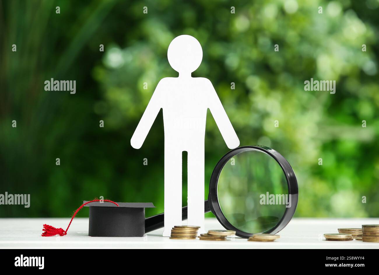 Human figure with magnifier, graduation hat and coins on table outdoors ...
