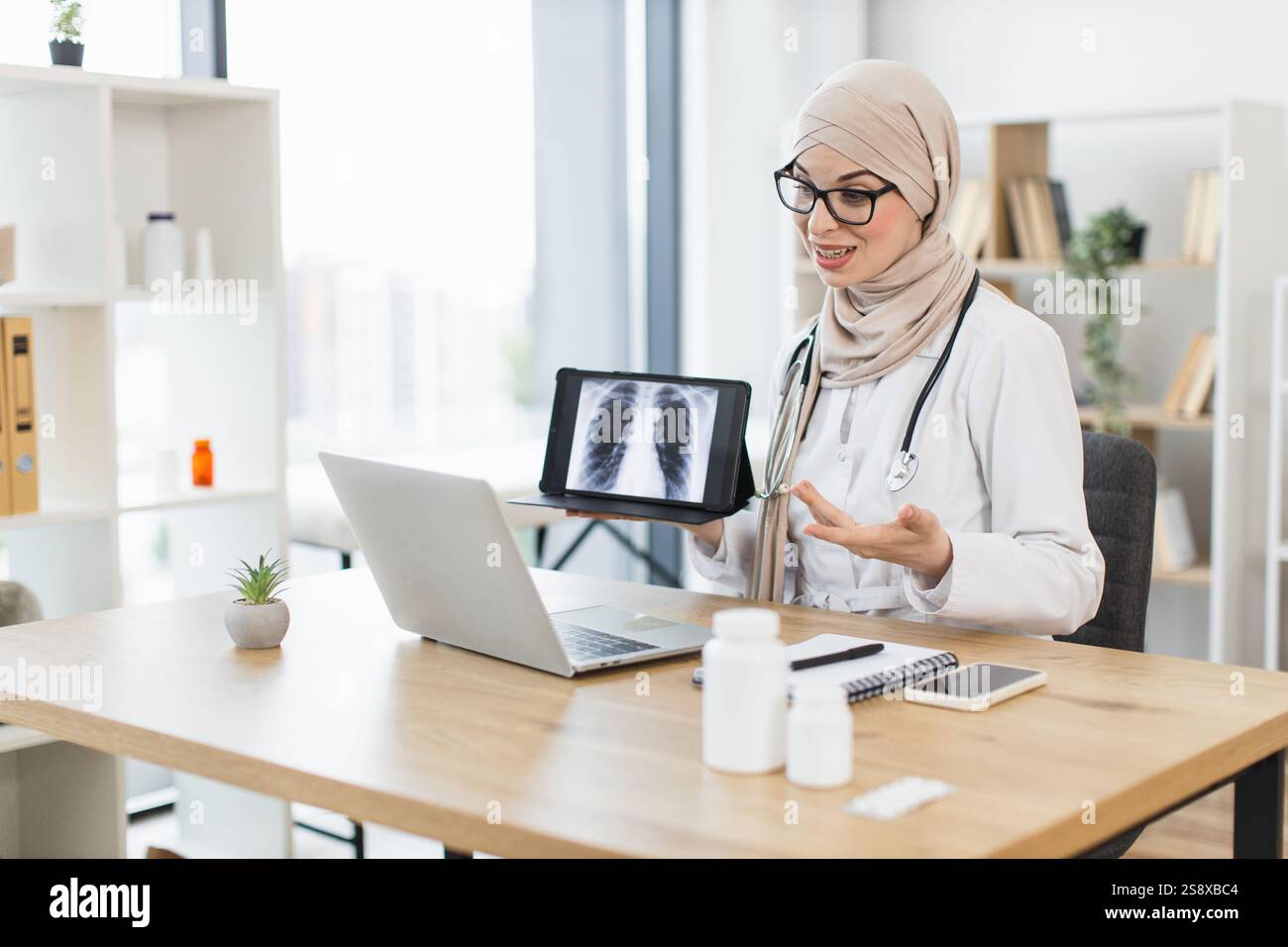 Muslim female doctor in hijab having video call sharing x-ray of diseased lung. Young adult in ...