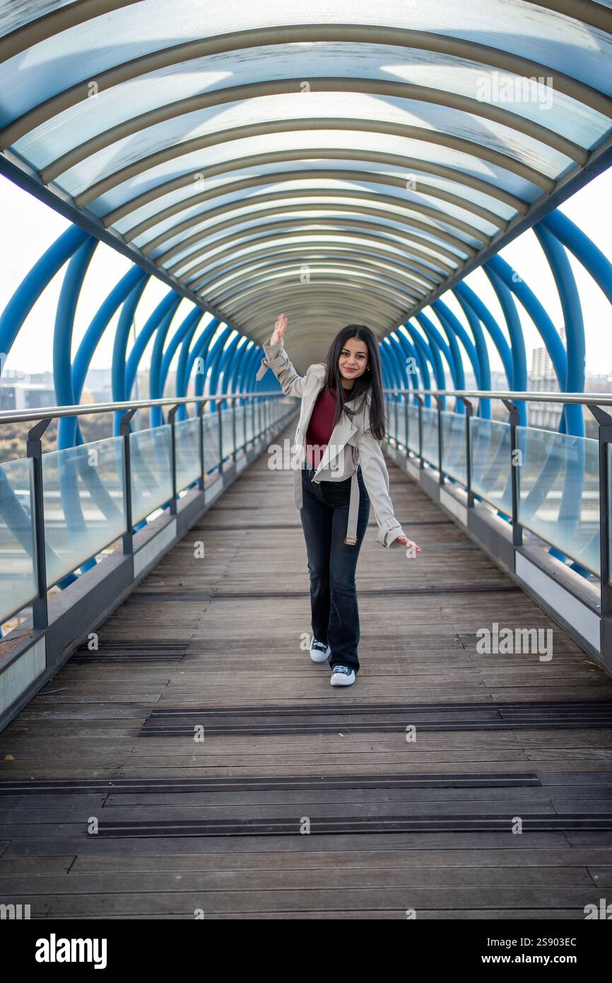 Stylish young woman waving while walking on a modern pedestrian bridge ...