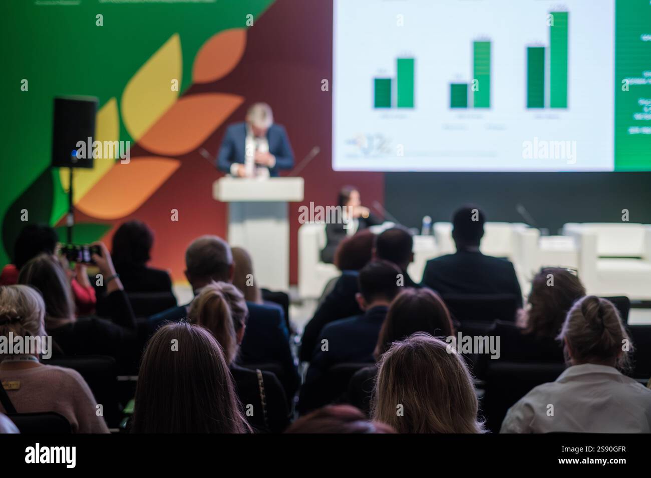 Speaker addresses audience at business conference with charts on screen ...