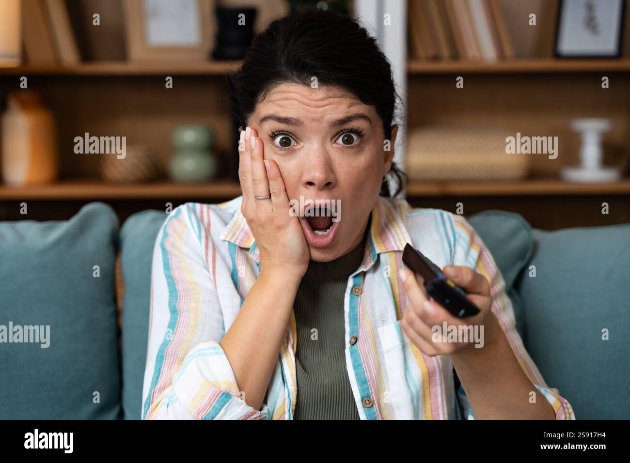 Facial expression. Young woman sitting alone at home on sofa, holding ...