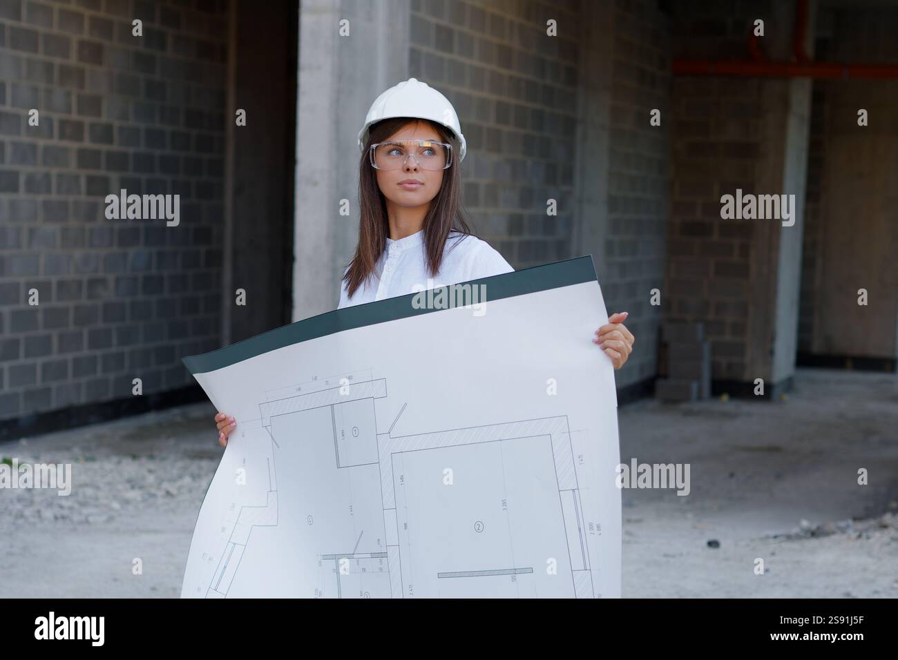 Young female architect wearing safety helmet and glasses, holding blueprint and inspecting ...
