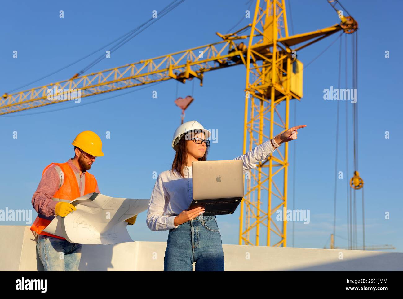 Construction engineers reviewing blueprints and using laptop on site with crane in background ...