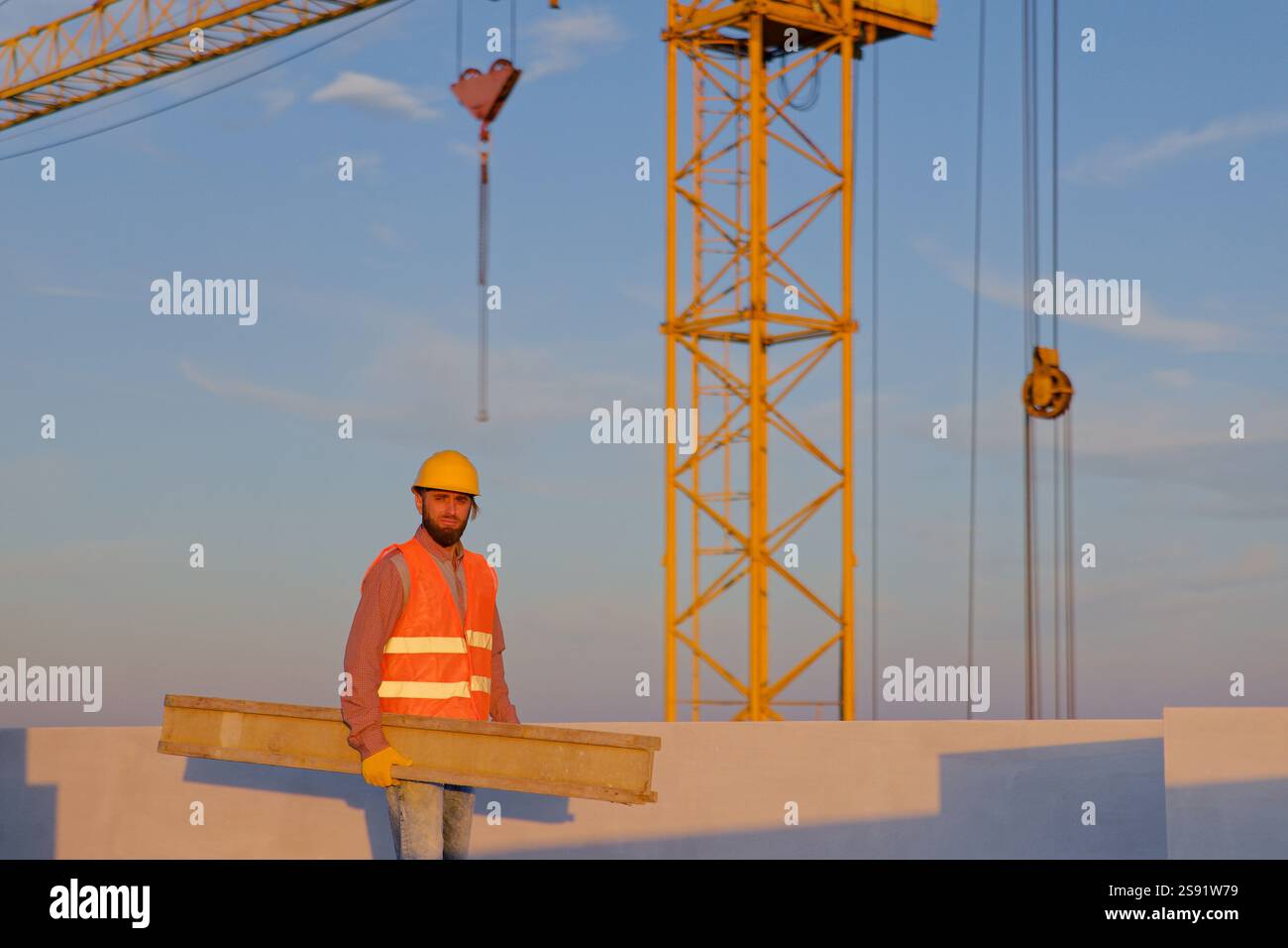 Construction worker carrying a wooden plank on a rooftop at sunset ...
