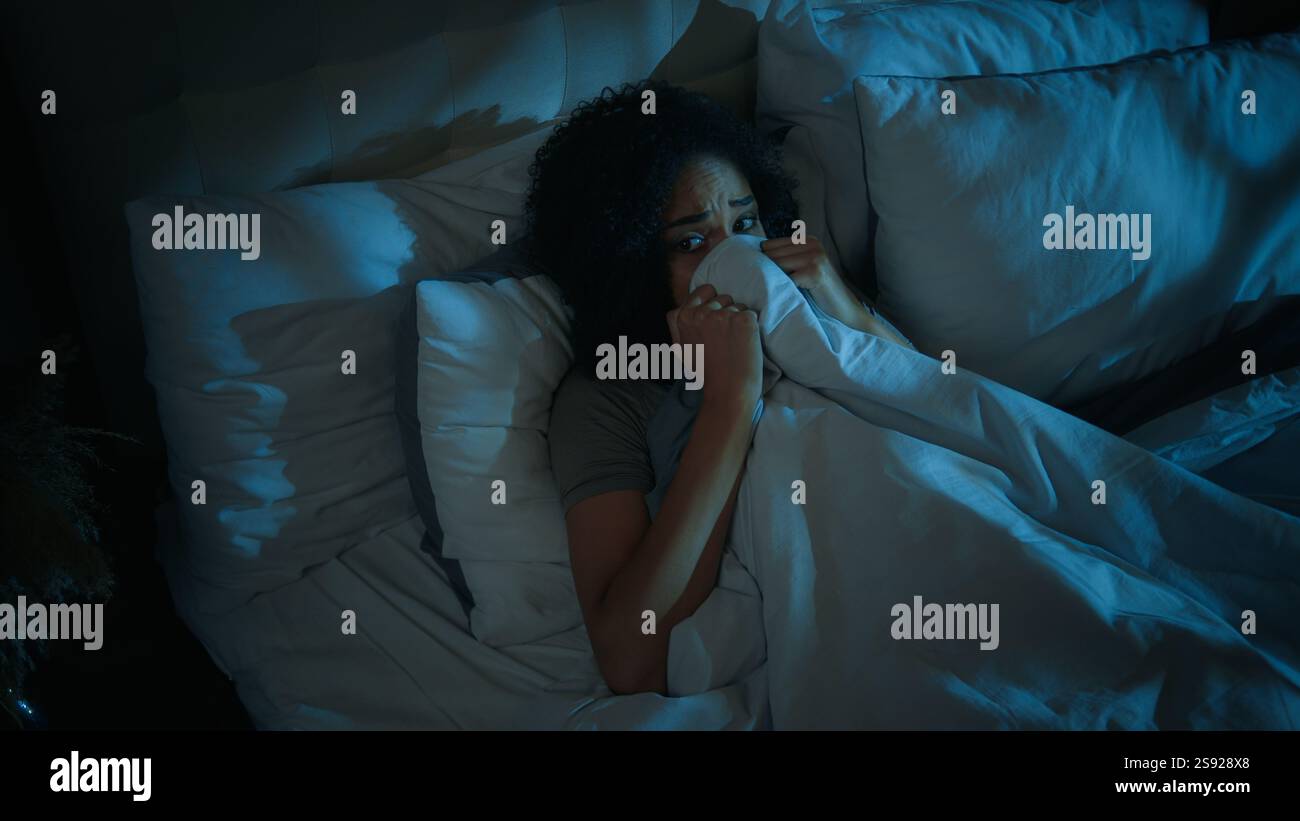 Scared frightened African American woman hiding under blanket bed nightmare sleep disorder bad ...