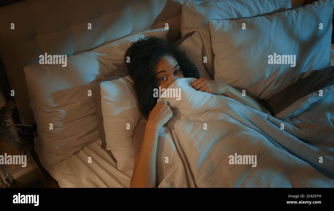 Scared frightened African American woman hiding under blanket bed nightmare sleep disorder bad ...