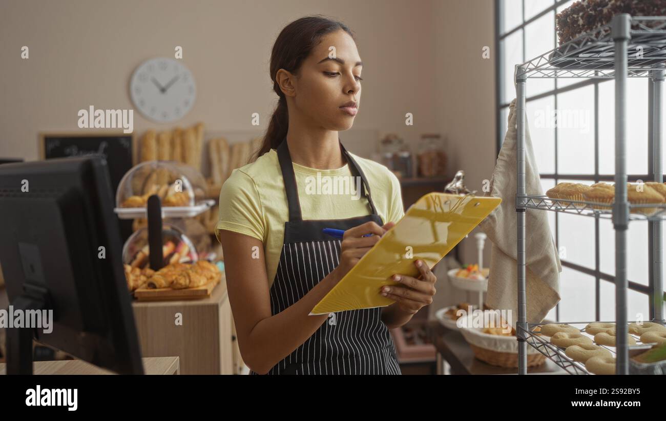 Young woman working in a bakery, checking inventory on a clipboard ...