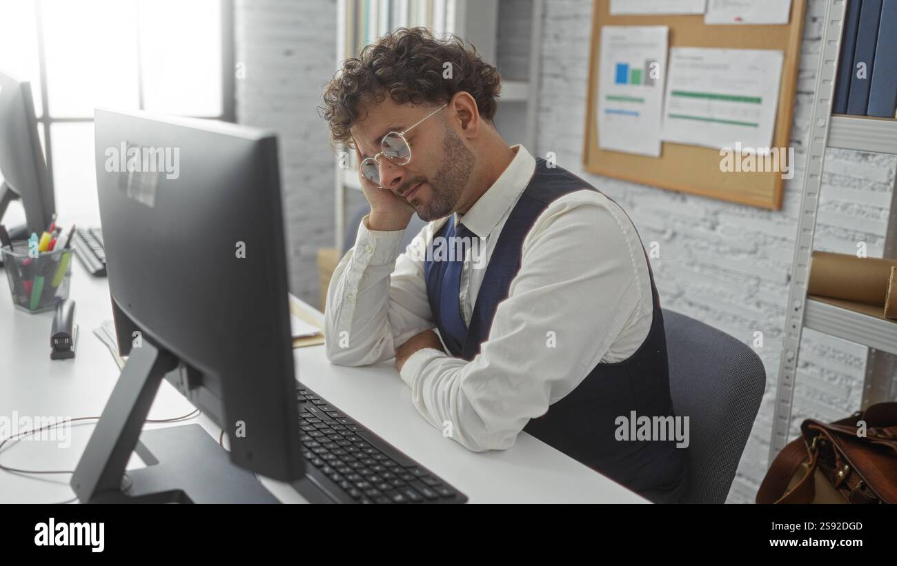Young man in an office looking tired and resting his head on his hand ...