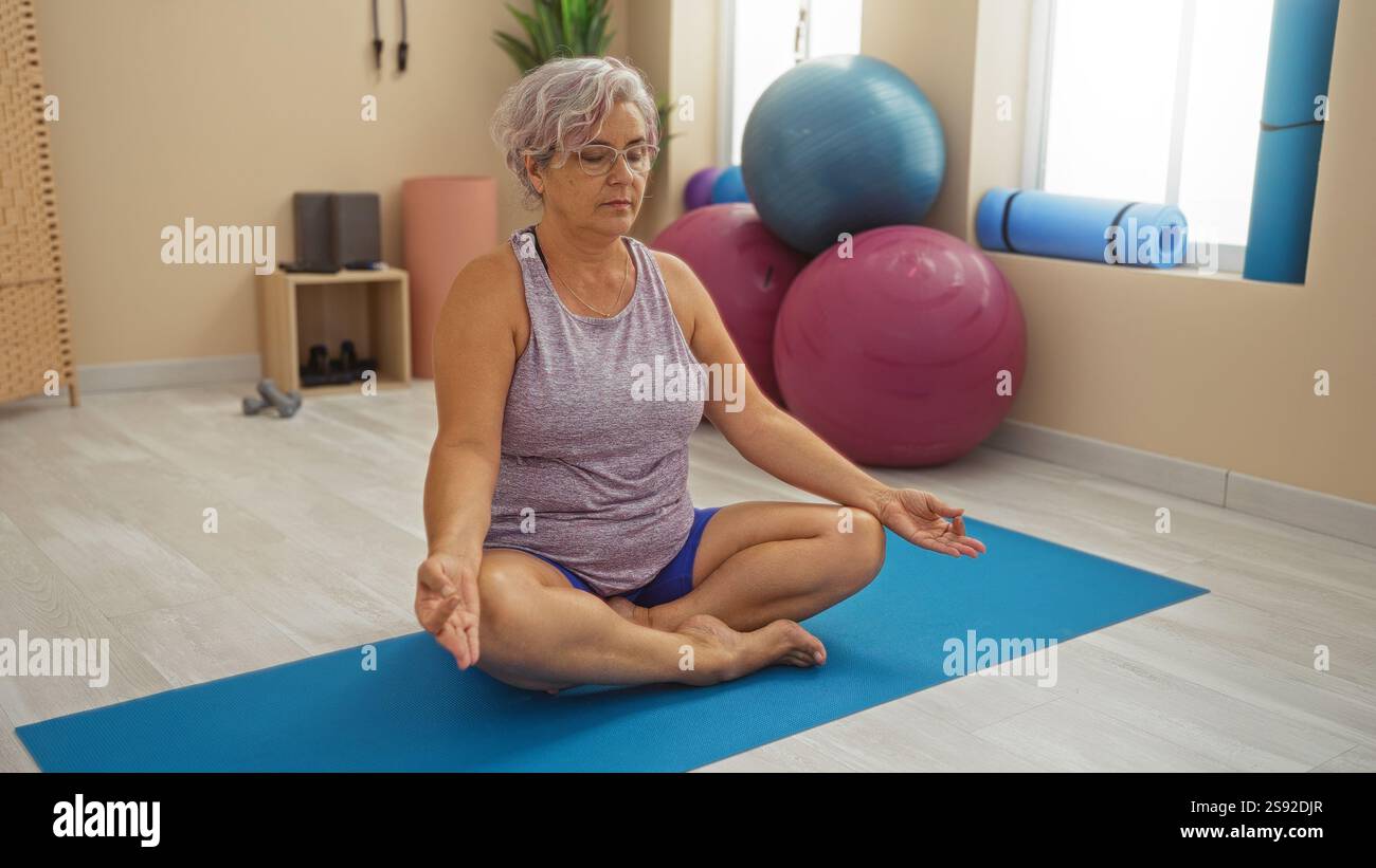 Middle-aged woman with grey hair meditating indoors on a yoga mat ...