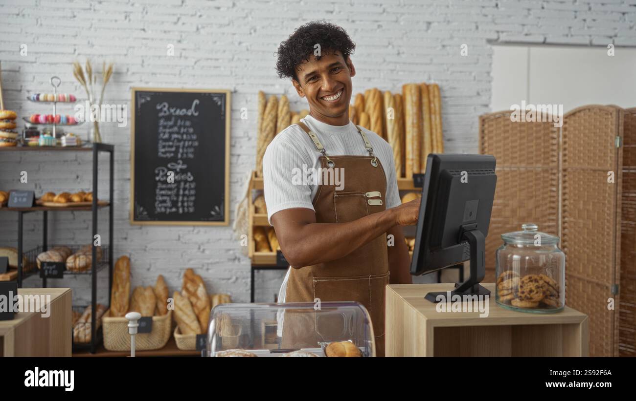 Young man working in a bakery shop, smiling while using a computer with ...