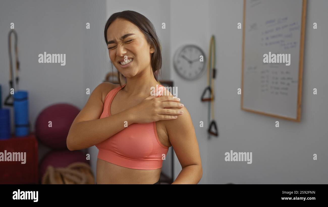 Young woman in a fitness center holding her shoulder in pain, indicating possible injury during ...