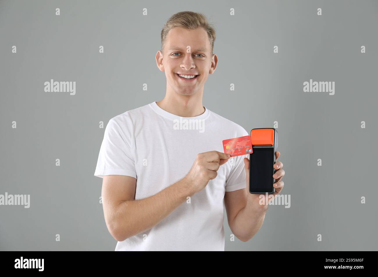 Happy young man with payment terminal and debit card on light grey ...