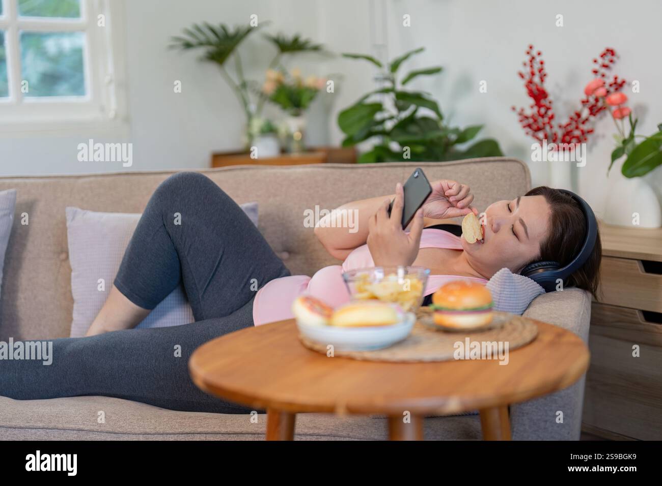 Overweight woman relaxing on the couch, enjoying snacks while scrolling ...