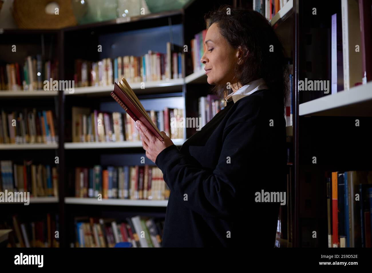 A contemplative woman standing by bookshelves in a library, reading a ...