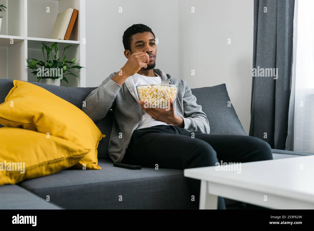 African american man focuses on the television while eating popcorn, fully immersed in show ...