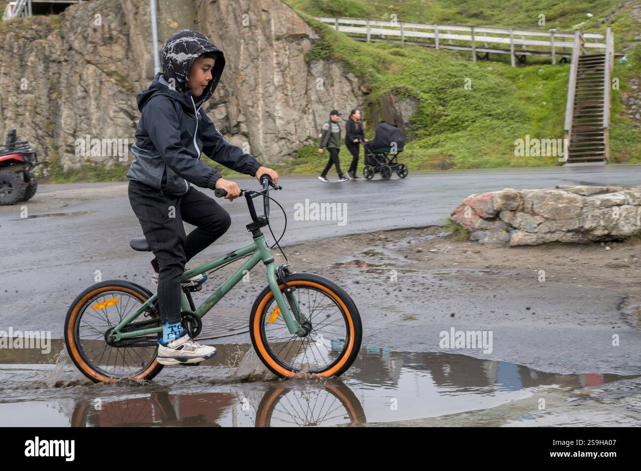 A child rides a bicycle through puddles on a wet roadway in Sisimiut ...