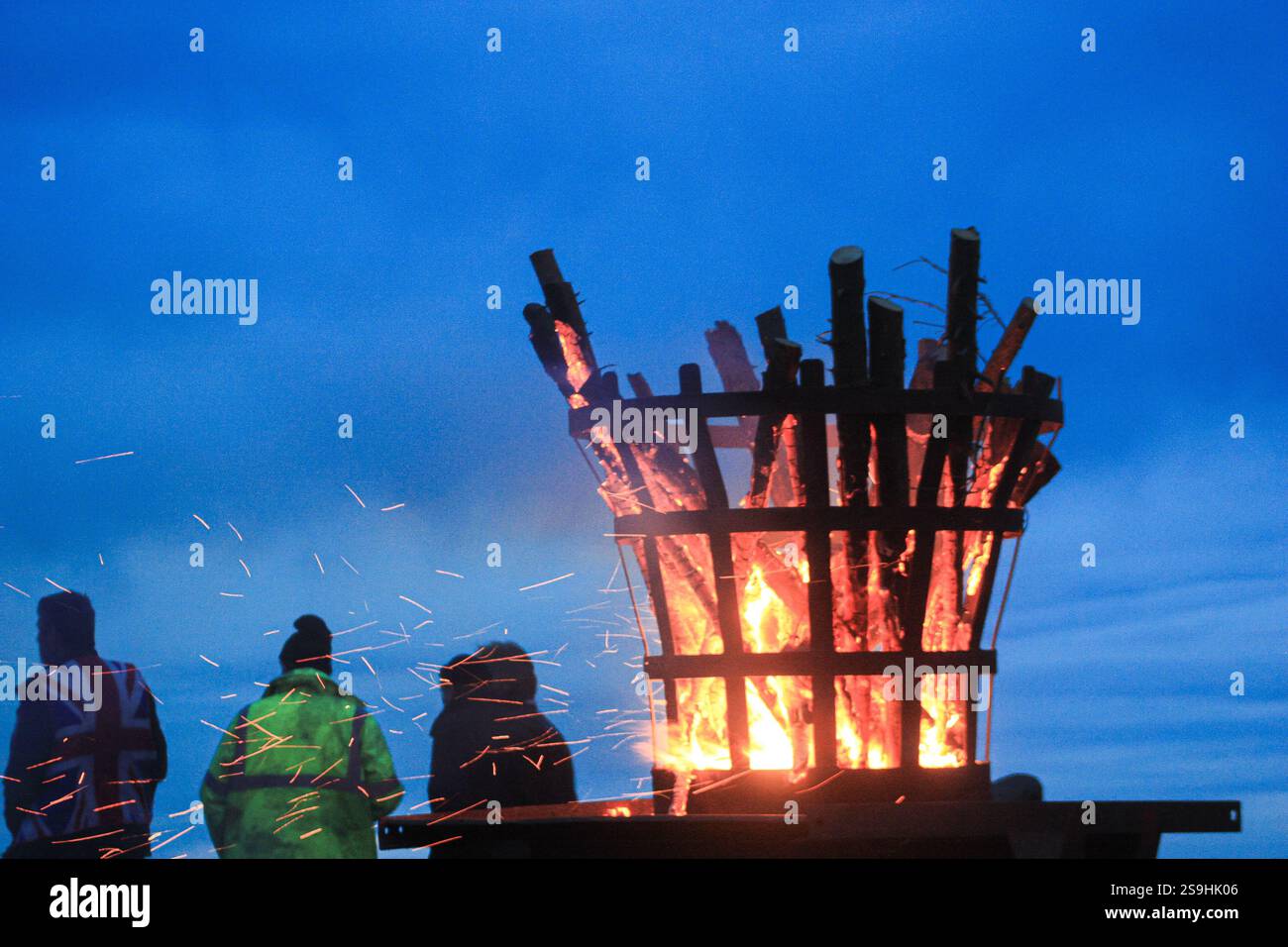 People gather around a fire beacon at dusk as sparks fly into the ...
