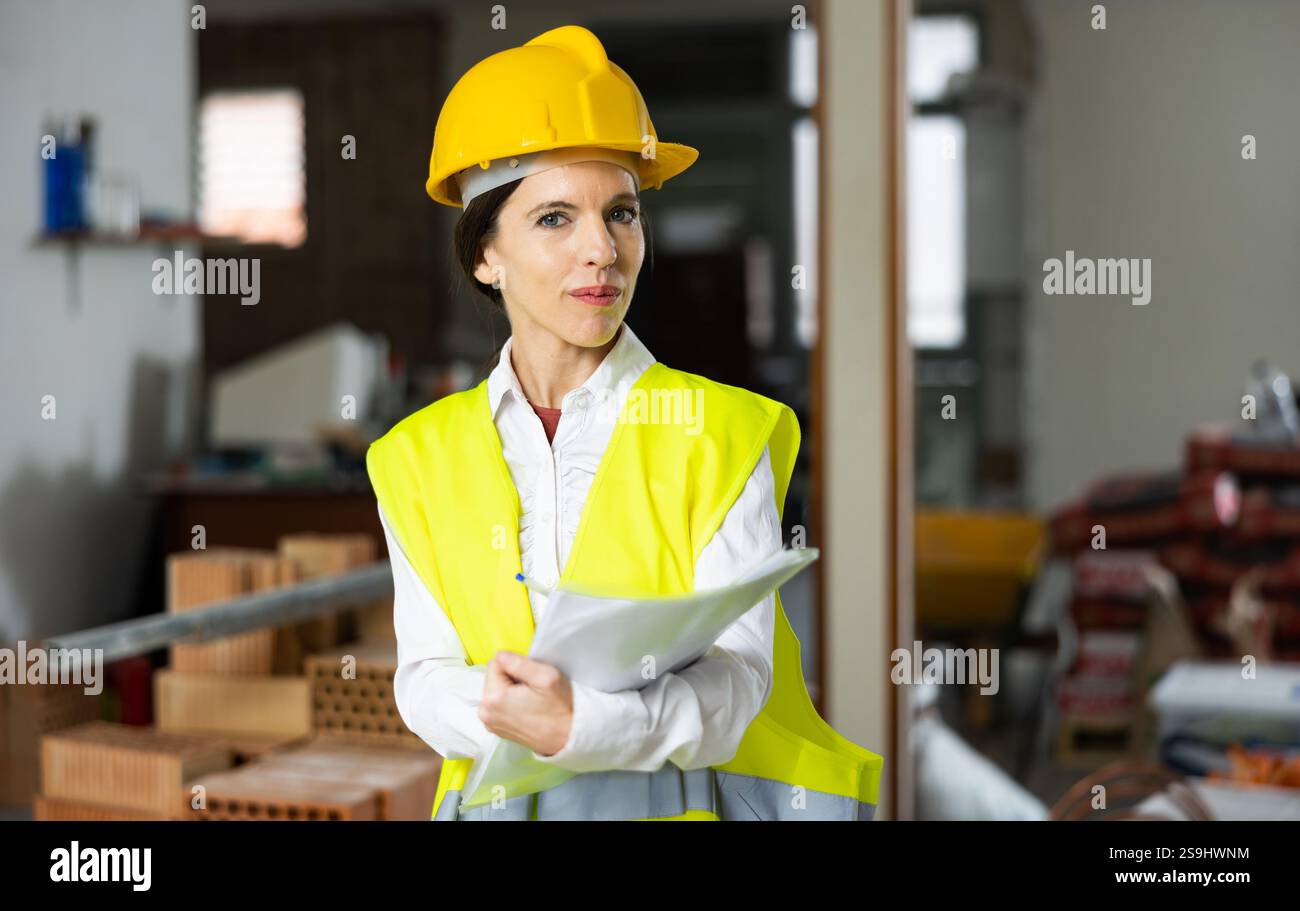 Portrait of woman builder in yellow vest and hardhat filling documentation during repair works ...