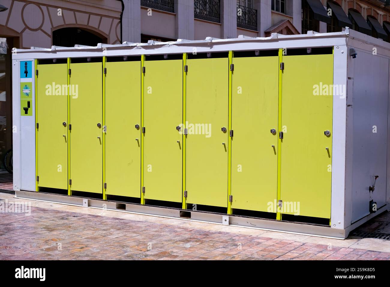 A portable, multi-stall, public restroom unit, painted in bright yellow and white, sits on a ...