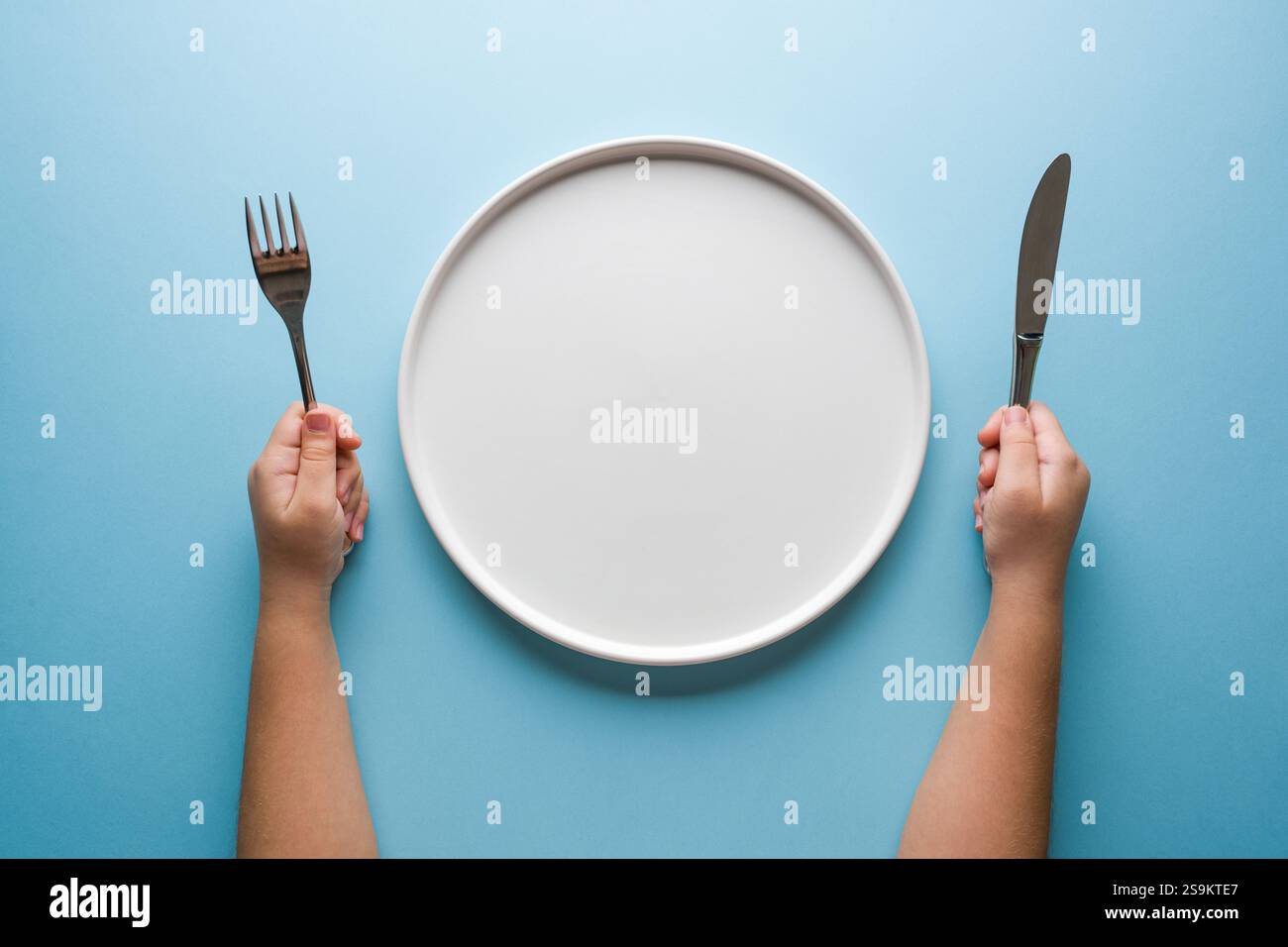Top view of girl's hands holding fork, knife and empty plate on blue ...