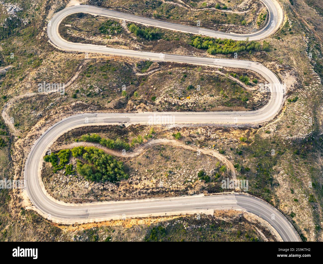 A winding asphalt road that passes through areas with few trees in ...