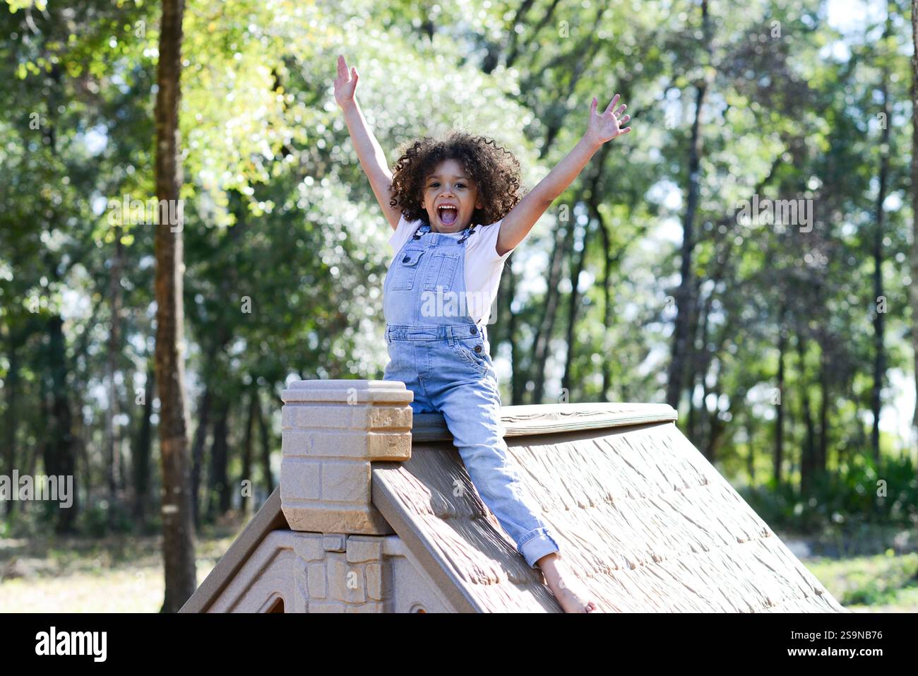A joyful child in overalls sits on a playhouse roof with arms ra Stock ...