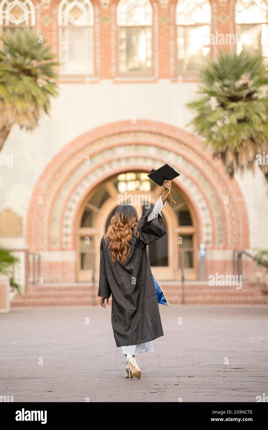 Woman graduate walking away holding up cap and wearing gown Stock Photo ...