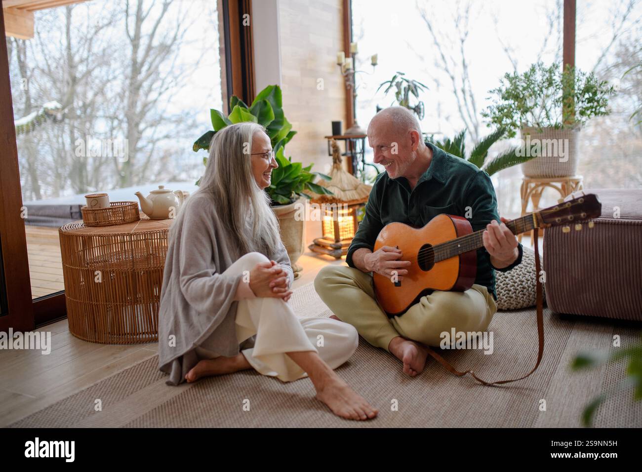 Real older couple at home. Husband playing music on a guitar for his ...