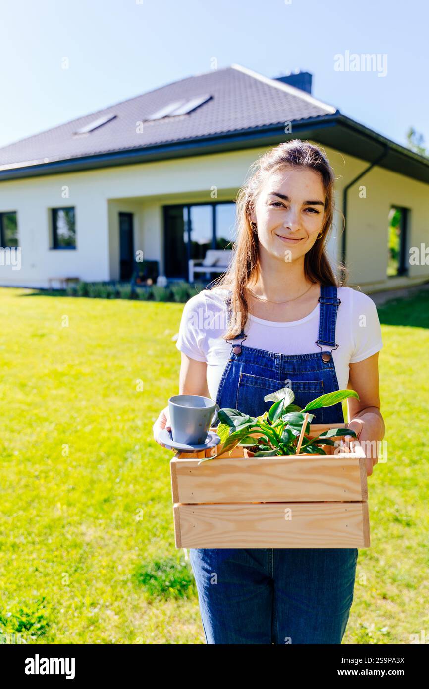 Young woman gardener wears jeans overalls standing in lawn holding box ...