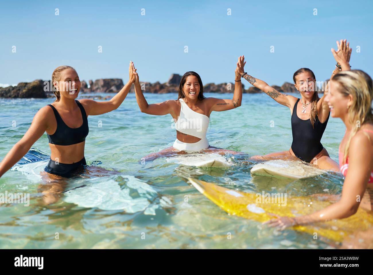 Four friends enjoy surfing in ocean. They sit on surfboards, high-five each other in shallow ...