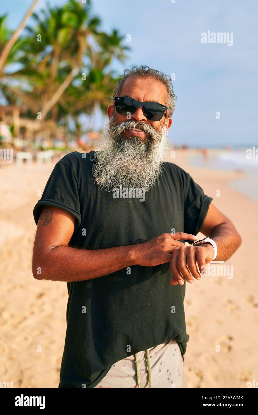 Senior bearded man with sunglasses on sandy beach uses smart watch ...