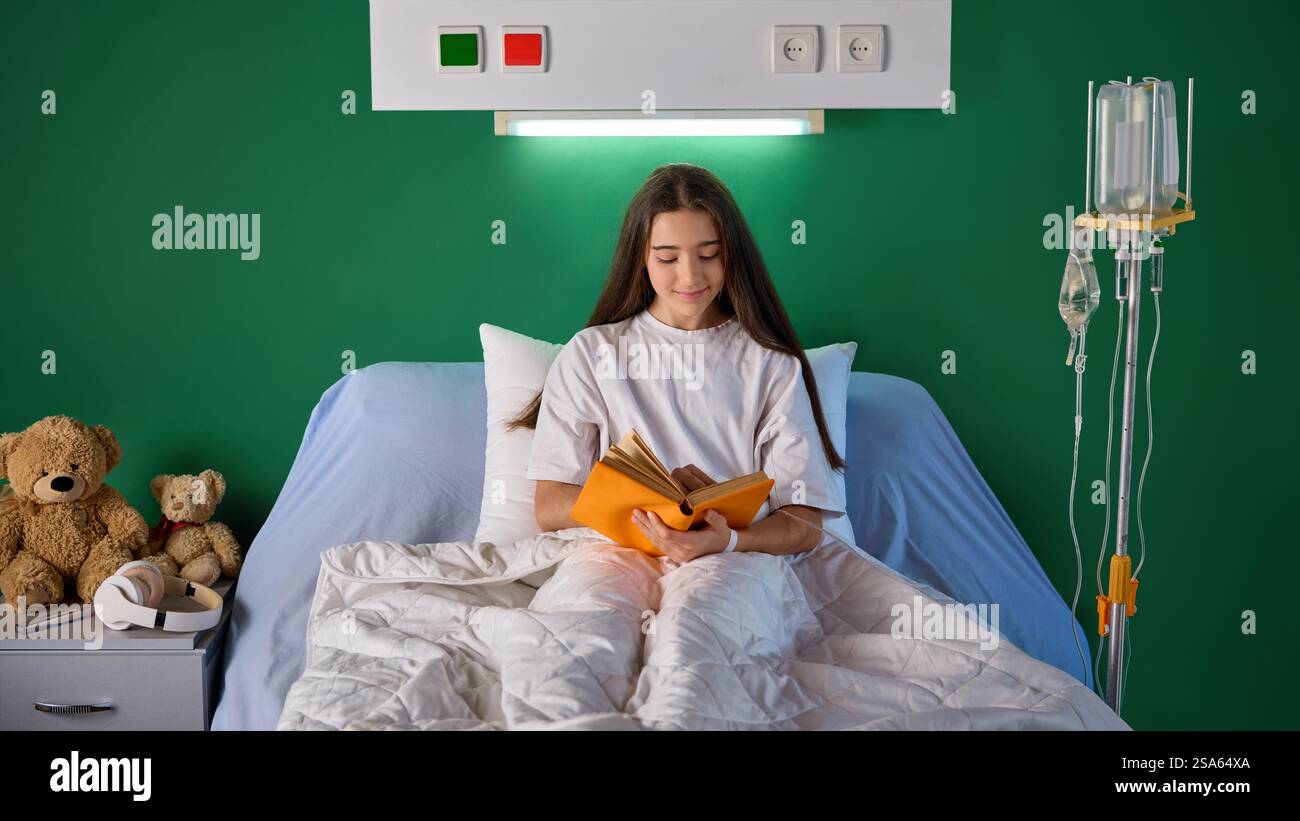 Teen girl in home wear sitting in bed in hospital ward and reading a ...