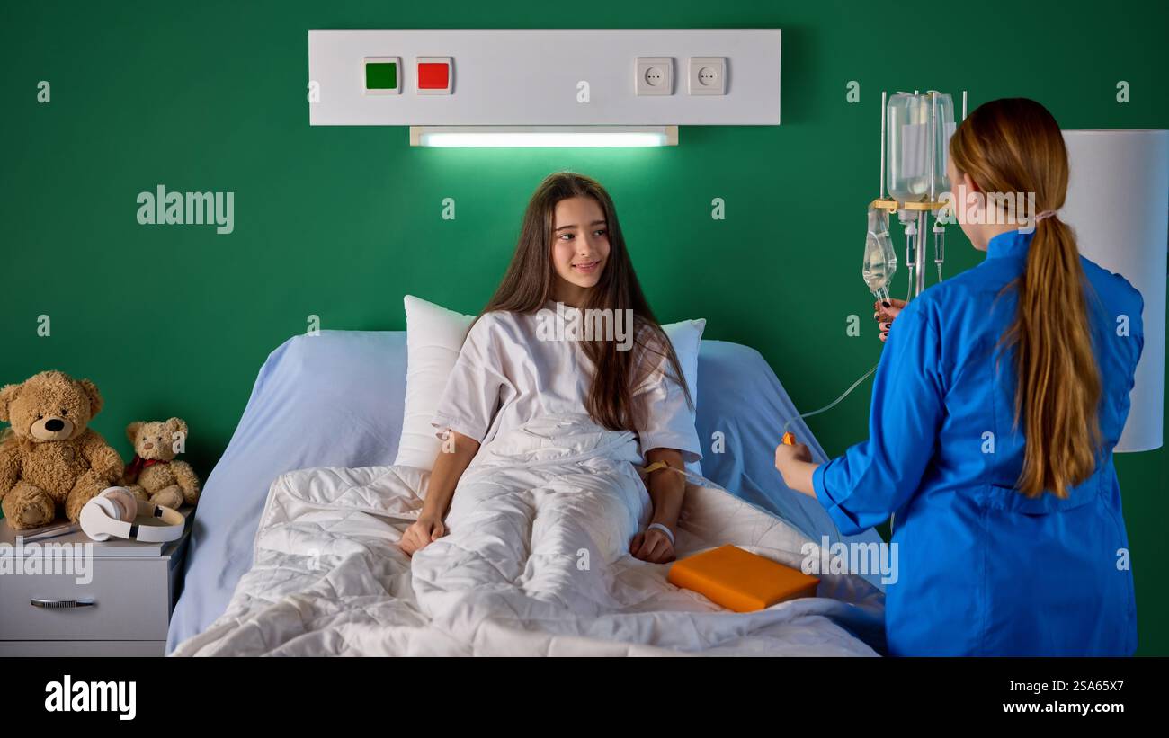 Teen girl sitting in bed in hospital ward while female nurse in blue ...