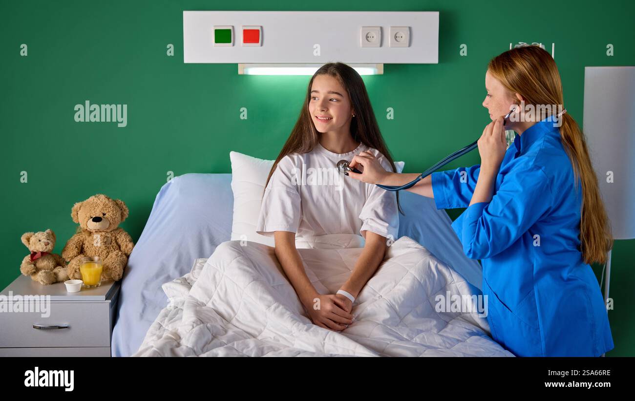 Teen girl sitting on bed in hospital ward while female nurse examining ...