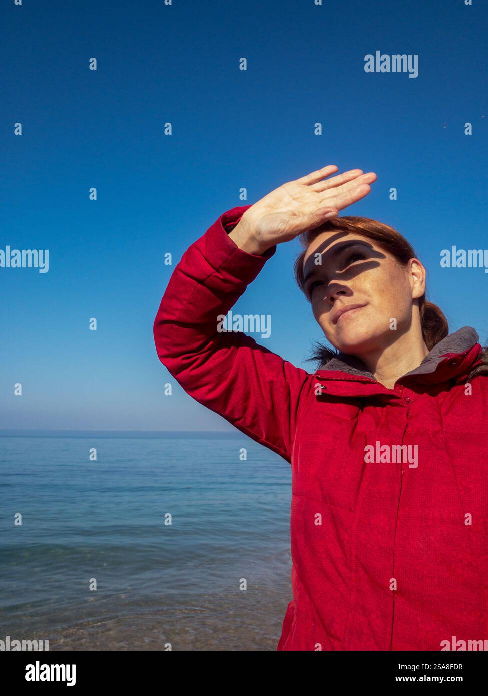 Woman in red puffer coat on sea beach looking up close her face by hand ...