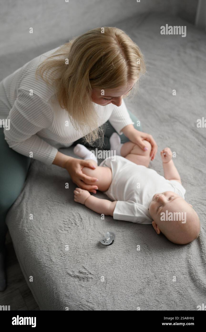 Woman does gymnastics to newborn baby lying on back on bed, spreading ...
